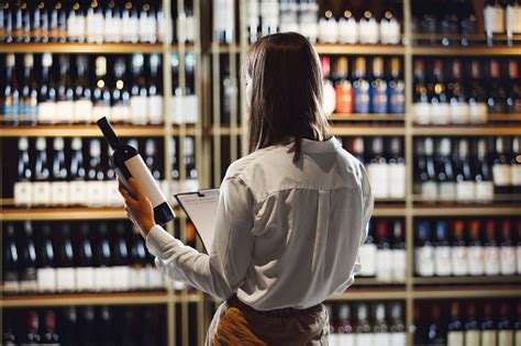 백인 젊은 여성 소믈리에가 선반에 다양한 레드 와인과 화이트 와인과 함께 와인 저장고 배경에 서 있는 레드 와인 한 병을 들고 있습니다 고름에 대한 스톡 사진 및 기타