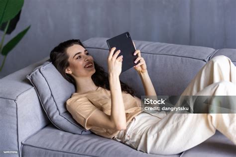 캐주얼 옷을 입은 아름다운 젊은 여성이 집에서 소파에 앉아 디지털 태블릿을 사용하고 있다 디지털 태블릿 사용하기에 대한 스톡 사진 및 기타 이미지 디지털 태블릿 사용하기