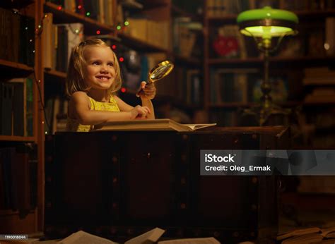 집 도서관에 있는 한 어린 소녀가 돋보기를 들고 있는 오래된 가슴에 앉아 있는 책을 읽고 있다 어린이와 교육 바닥에 많은 책 야간 가정 생활에 대한 스톡 사진 및 기타