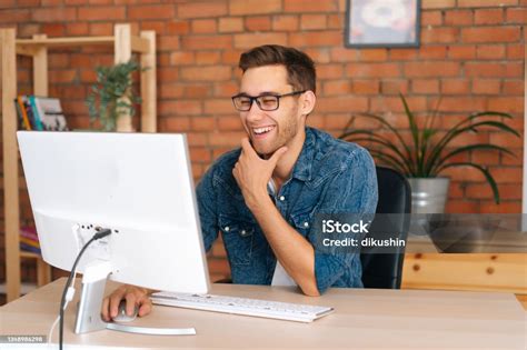 현대적인 인테리어가 있는 집 사무실 책상에 앉아 있는 데스크탑 컴퓨터에서 일하는 세련된 안경을 쓰고 웃는 젊은이의 중간 샷