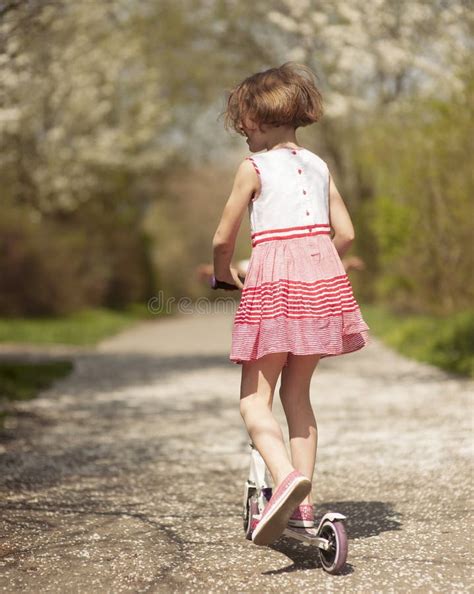 Young Girl Riding Scooter Away From Camera On Path Through Park Stock