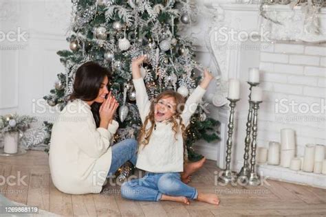 새해 전야에 행복한 가족 젊은 아름다운 어머니와 흰색 아늑한 스웨터와 담요에 작은 귀여운 딸은 장식 된 크리스마스 트리 아래에서 재미와 포옹을합니다 축제 홈 클래식 인테리어