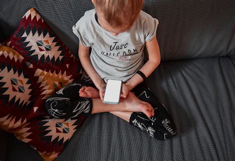 거실의 소파에 앉아있는 귀여운 아이 소녀는 스마트 폰을 사용하고 온라인 게임을합니다 전화를 들고 화면 장치를보고 아이는 소파에 앉아서 거실에서 휴식을 취합니다 기술 개념