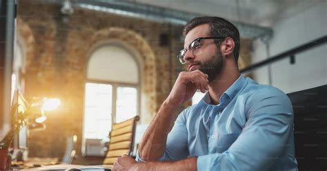 생각 중 안경을 쓰고 정장을 입은 젊은 수염 난 남자가 컴퓨터로 작업하고 현대 사무실에 앉아 턱을 만지고 있습니다 사업 개념입니다 작업 개념입니다 현대 기술 사진