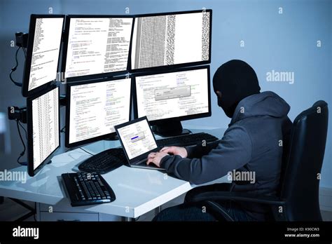Male Hacker Using Laptop Against Multiple Monitors At Desk In Office