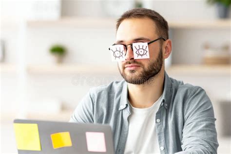 Tired Millennial Guy Having Stickers With Drawings Of Open Eyes On Glasses Sleeping In Front Of