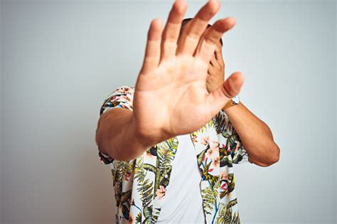 휴일에 젊은 잘생긴 남자는 손으로 눈을 덮고 슬프고 두려움 표정으로 중지 제스처를하고 흰색 배경 위에 하와이 셔츠를 입고 부끄럽고 부정적인 개념 남자에 대한 스톡 사진 및