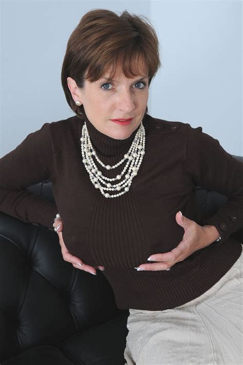 A Woman Sitting In A Chair With Her Hands On Her Hips Wearing Pearls And A Brown Sweater