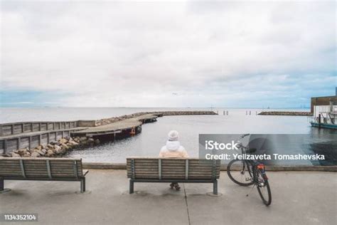 젊은 백인 여자는 흐린 날씨에 겨울에 코펜하겐 덴마크의 바닷가에 발트 해를 내려다 볼 수 있는 나무 벤치에 그녀의 뒤에 앉아 인근에 있는 소녀 산책 곤 오스카 자전거 가을에