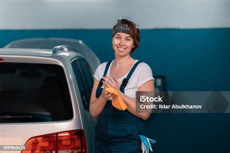 푸른 작업복을 입은 행복한 백인 젊은 여성이 헝겊으로 손을 흔들고 있는 모습 배경에있는 자동차와 워크샵 자동차 수리점에서 일하기 기계 부분에 대한 스톡 사진 및 기타 이미지