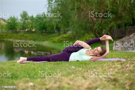 얇은 갈색 머리 소녀는 스포츠를 하고 여름 공원에서 아름답고 세련된 요가 포즈를 취합니다 녹색 무성한 숲과 배경에 강 요가 매트에서 운동을하는 여자 가냘픈에 대한 스톡 사진