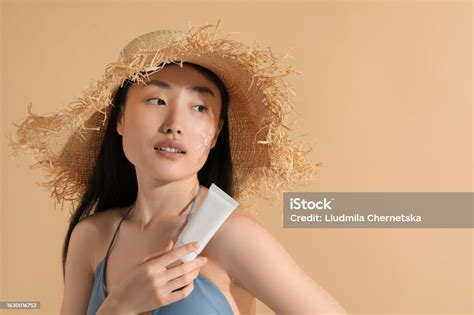 얼굴에 자외선 차단제를 바르고 있는 밀짚모자를 쓴 아름다운 젊은 여성이 베이지색 배경에 자외선 차단 크림을 들고 있다 햇볕 차단 로션에 대한 스톡 사진 및 기타 이미지