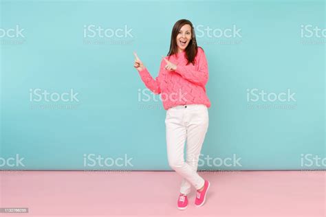 니트 로즈 스웨터 스튜디오에서 밝은 분홍색 파란색 파스텔 벽 배경에 고립 된 포즈 흰색 바지에 웃는 젊은 여자의 전체 길이 초상화 패션 라이프 스타일 개념입니다 복사 공간을