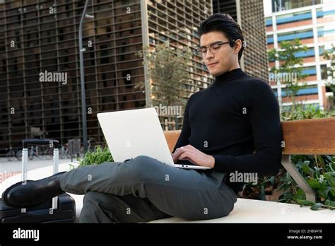Serious Asian Male Entrepreneur Typing On Netbook While Sitting On Border Near Suitcase On