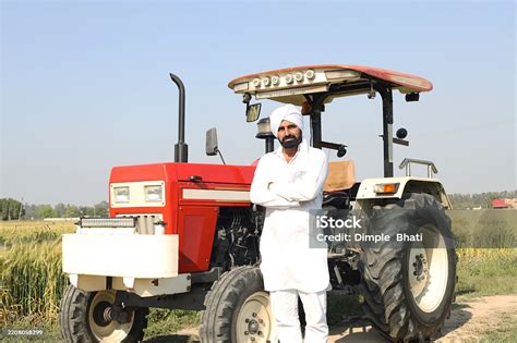 농업 현장에서 트랙터와 함께 서 있는 하리아나 농부의 펀자브 흰색 전통 쿠르타와 터번을 쓴 인도 시크교도 펀잡 남자가 카메라를 흰색으로 보고 팔짱을 끼고 있습니다 인도