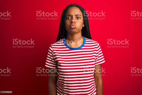 젊은 아프리카 계 미국인 여자 스트라이프 티셔츠 를 입고 고립 된 빨간 배경 퍼핑 뺨 위에 서 재미있는 얼굴 공기 미친 표정으로 입이 팽창 땋은 머리에 대한 스톡 사진 및