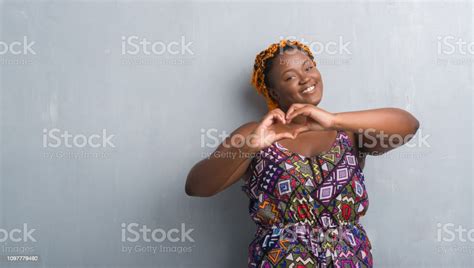 심장 기호와 손으로 모양을 보여주는 사랑에 웃 고 오렌지 머리 띠를 입고 회색 그런 지 벽에 젊은 아프리카계 미국인 여자 낭만적인 개념입니다 땋은 머리에 대한 스톡 사진 및