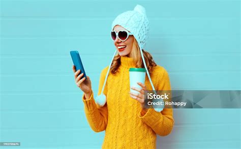 노란색 니트 스웨터를 입고 전화와 커피 컵을 입고 젊은 여자를 미소 겨울 초상화 Pom Pom와 흰색 모자 파란색 배경에 하트 모양선글라스 겨울에 대한 스톡 사진 및 기타