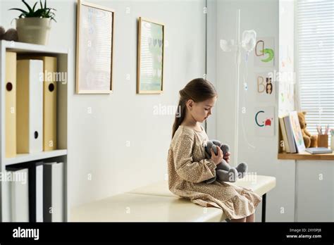 Young Girl Sitting On Bench In Pediatric Clinic Holding Stuffed Toy With Educational Posters