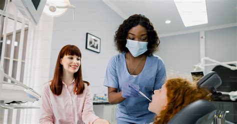 두려움없는 치과 치료 파란색 유니폼과 마스크를 쓴 아프리카 여성 치과 의사가 현대 소아 치과 진료소에서 어머니의 손을 잡고 어린 소녀의 치아를 검사합니다 사진