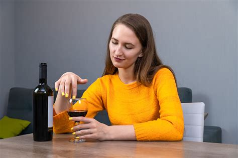 오렌지색 스웨트셔츠를 입은 젊은 검은 머리의 여성이 부엌 테이블에 앉아 어두운 유리잔을 들고 와인 한 병 옆에 빨간 와인 한 잔을 들고 손가락을 대고 있다 가정 생활에 대한