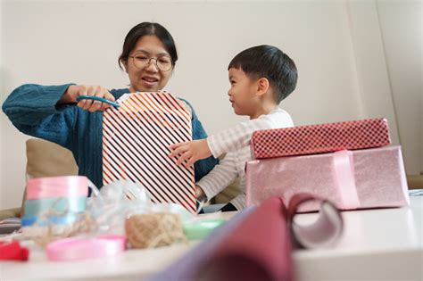 즐거운 아시아 아이가 부모를 도와 테이블 위에 놓인 형형색색의 선물을 풀어줍니다 방은 밝고 분위기는 축제 분위기로 행복한 휴가 모임을 반영합니다 2명에 대한 스톡 사진 및