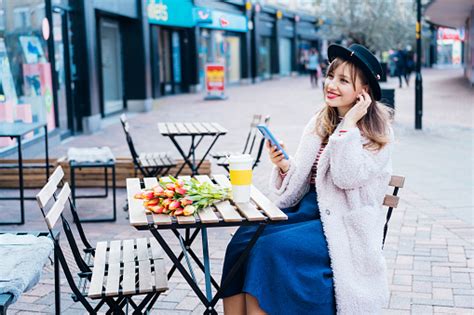 재사용 가능한 컵에서 커피를 즐기고 음악을 듣고 무선 이어폰을 사용하는 팟캐스트와 거리 카페에서 전화를 하는 세련된 젊은 미소 짓는 여성 테이블에 신선한 튤립 꽃다발 봄철