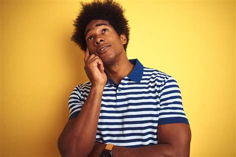 해군 줄무늬 폴로를 입고 고립 된 노란색 배경 위에 서있는 아프로 머리를 가진 미국 남자는 턱에 손을 얹고 질문 펜시브 표현을 생각하고 있습니다 사려 깊은 얼굴로 미소 의심