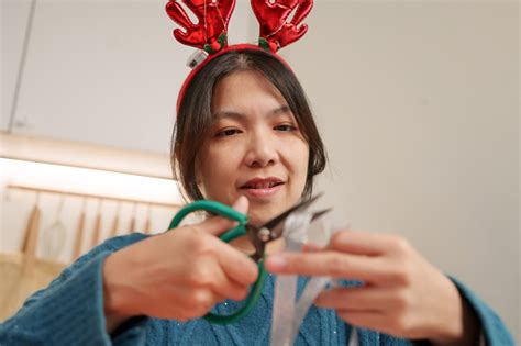 순록 뿔을 가진 아시아 여성은 아늑한 실내 환경에서 휴일 공예품을 작업하면서 가위로 리본 조각을 조심스럽게 자릅니다 분위기는 축제적이고 즐겁습니다 40 44세에 대한 스톡