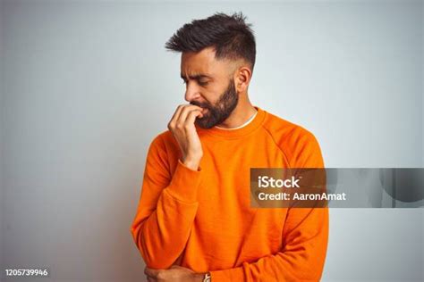 고립 된 흰색 배경 위에 오렌지 스웨터를 입은 젊은 인도 남자는 손톱을 물고 손톱을 물고 손에 스트레스와 긴장 찾고 불안 문제 남자에 대한 스톡 사진 및 기타 이미지