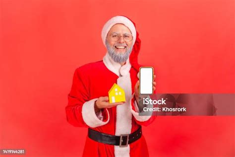 회색 수염을 가진 웃는 노인의 초상화는 작은 종이 집과 스마트 폰 위트 빈 화면을 들고 산타 클로스 의상을 입고 있습니다 30 34세에 대한 스톡 사진 및 기타 이미지