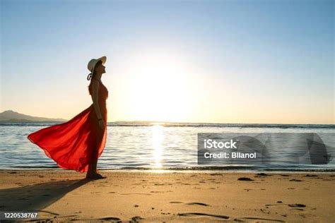 긴 빨간 드레스와 밀짚 모자를 쓰고 있는 젊은 여성이 초여름 아침에 떠오르는 태양의 경치를 즐기는 바닷가의 모래 해변에 서 있습니다 드레스에 대한 스톡 사진 및 기타 이미지