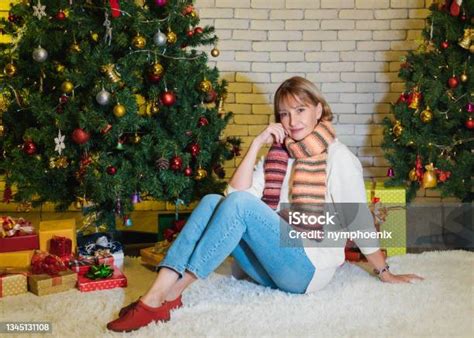 아름다운 선임 백인 여성은 거실에서 선물이있는 장식 된 크리스마스 트리 앞에서 흰색 모피 카펫에 혼자 앉아 있는 것처럼 보입니다 50 54세에 대한 스톡 사진 및 기타
