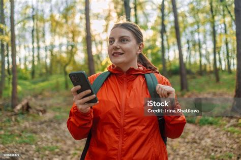 배낭을 메고 따뜻한 옷을 입은 젊은 여성 여행자가 봄 숲에 서서 전화기 화면을 바라보고 있다 등산로에 있는 여자들 숲에 있는 나무 길 폴란드 유럽의 국립 공원 고화질 사진