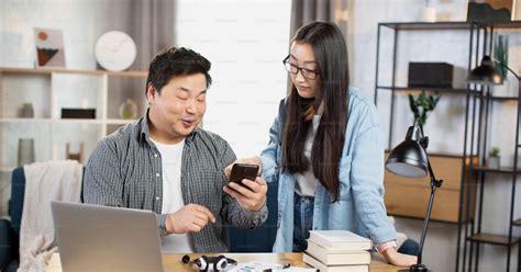 테이블에 앉아 스마트폰을 사용하는 아시아 남자 근처에 서 있는 아름다운 아시아 여성 캐주얼 복장을 한 두 명의 사무원 Qa 엔지니어 현대 기술로 작업하십시오 온라인