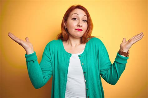 Youg 아름다운 빨간 머리 여자 겨울 녹색 스웨터 위에 고립 된 노란색 배경 단서와 팔과 손을 제기 와 혼란 표정 의심 개념 감정에 대한 스톡 사진 및 기타 이미지