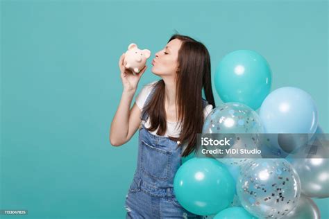 부드러운 젊은 여성이 손에 돼지 저금통에 공기 키스를 보내는 불어 축 하 파란색 청록색 배경에 고립 된 다채로운 공기 풍선을