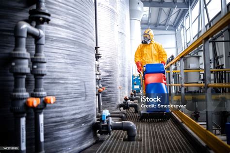 보호복을 입고 가스 마스크를 쓴 공장 노동자가 생산 공장 내부의 대형 산 저장 탱크로 화학 물질을 운반하는 카트를 밀고 있습니다 공장 산업 건물에 대한 스톡 사진 및 기타