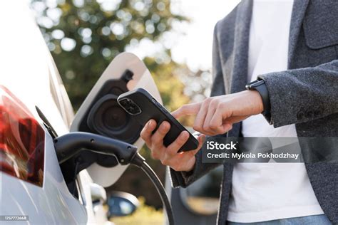 스마트폰 앱을 사용하여 충전소에서 전기 자동차를 충전하는 남성 충전소에서 전기 자동차의 플러그를 뽑는 알아볼 수 없는 남성 해질녘 전기 자동차의 전원 코드를 뽑고 있는 남자