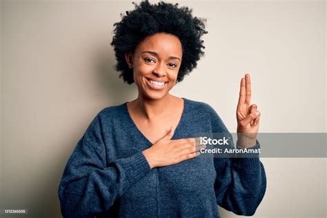가슴과 손가락에 손을 들고 맹세하는 캐주얼 스웨터를 입고 곱슬 머리를 한 젊은 아름다운 아프리카 계 미국인 아프로 여성이 충성약속 맹세를 합니다 만들기에 대한 스톡 사진 및