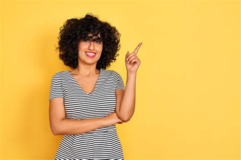 곱슬 머리를 가진 젊은 아랍 여성은 얼굴에 큰 미소와 고립 된 노란색 배경 위에 줄무늬 드레스를 입고 카메라를보고 측면에 손과