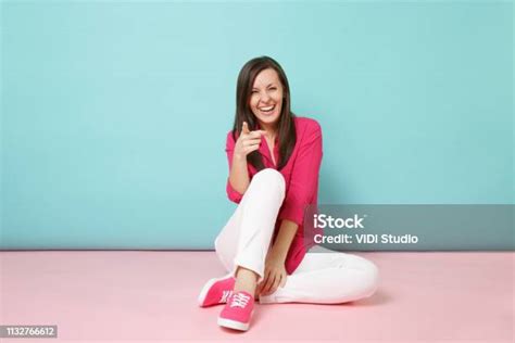 장미 셔츠 블라우스 밝은 분홍색 파란 파스텔 벽 배경 스튜디오에 고립 된 바닥에 앉아 흰색 바지에 웃는 젊은 여자의 전체 길이