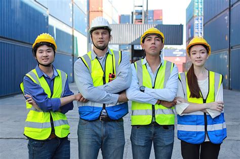 안전모를 쓰고 팔짱을 낀 보호복을 입은 다민족 기술자 엔지니어 및 사업가 그룹이 컨테이너 화물 현장에서 함께 성공하거나 거래를