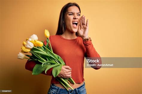 젊은 아름 다운 갈색 머리 여자 노란색 배경 에 튤립 꽃다발 을 들고 외치고 큰 소리로 옆으로 입에 손으로 비명 통신 개념입니다 갈색 머리에 대한 스톡 사진 및 기타 이미지