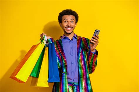 밝은 노란색 배경에 스마트 폰으로 컬러 쇼핑백을 들고있는 활기찬 옷차림의 쾌활한 젊은이입니다 20 24세에 대한 스톡 사진 및 기타 이미지 Istock