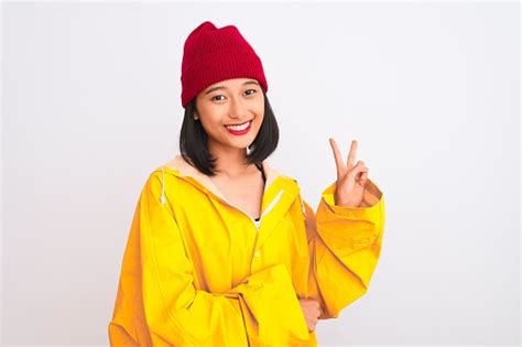 비옷과 울 모자를 쓰고 고립된 흰색 배경 위에 행복한 얼굴을 하고 승리의 표지판을 들고 있는 젊은 아름다운 중국 여성 2번 보여주기에 대한 스톡 사진 및 기타 이미지