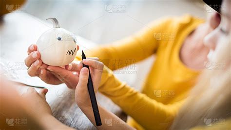 可爱的小女孩坐在桌子旁，和她的妈妈一起装饰白色的万圣节南瓜。手的细节。万圣节和家庭生活方式背景。