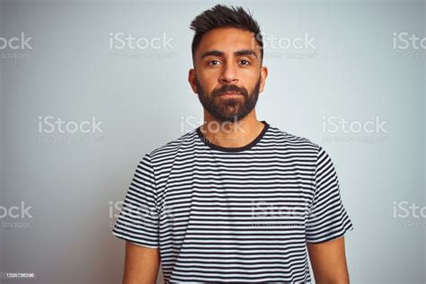 얼굴에 심각한 표정으로 고립 된 흰색 배경 위에 서있는 검은 줄무늬 티셔츠를 입은 젊은 인도 남자 카메라를 보는 간단하고 자연스러운 T 셔츠에 대한 스톡 사진 및 기타