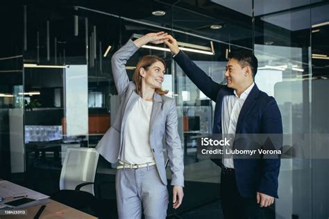 두 명의 직원이 프로젝트의 성공적인 납품을 축하하며 아시아 남자와 금발의 여성이 현대적인 사무실에서 기뻐하고 행복한 동료를 함께 춤을 추는 것을 축하합니다 사무실에 대한