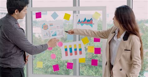 비즈니스 사람들은 사무실의 프로젝트 계획 보드에서 일하고 동료 친구와 대화하여 프로젝트 개발을 분석합니다 그들은 유리 벽에 게시 된 스티커 메모를 사용하여 정리합니다